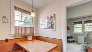 Uma área de refeições com um banco e mesa de madeira, uma janela com persianas e um quadro pendurado na parede.