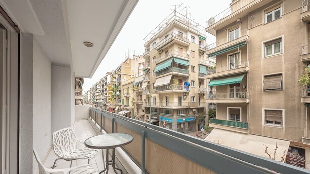 Varanda com cadeiras brancas e uma pequena mesa, com vista para uma fileira de edifícios altos com varandas e toldos.
