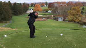 Um campo de golfe com um jogador em pleno movimento, um lago e árvores de outono ao fundo.