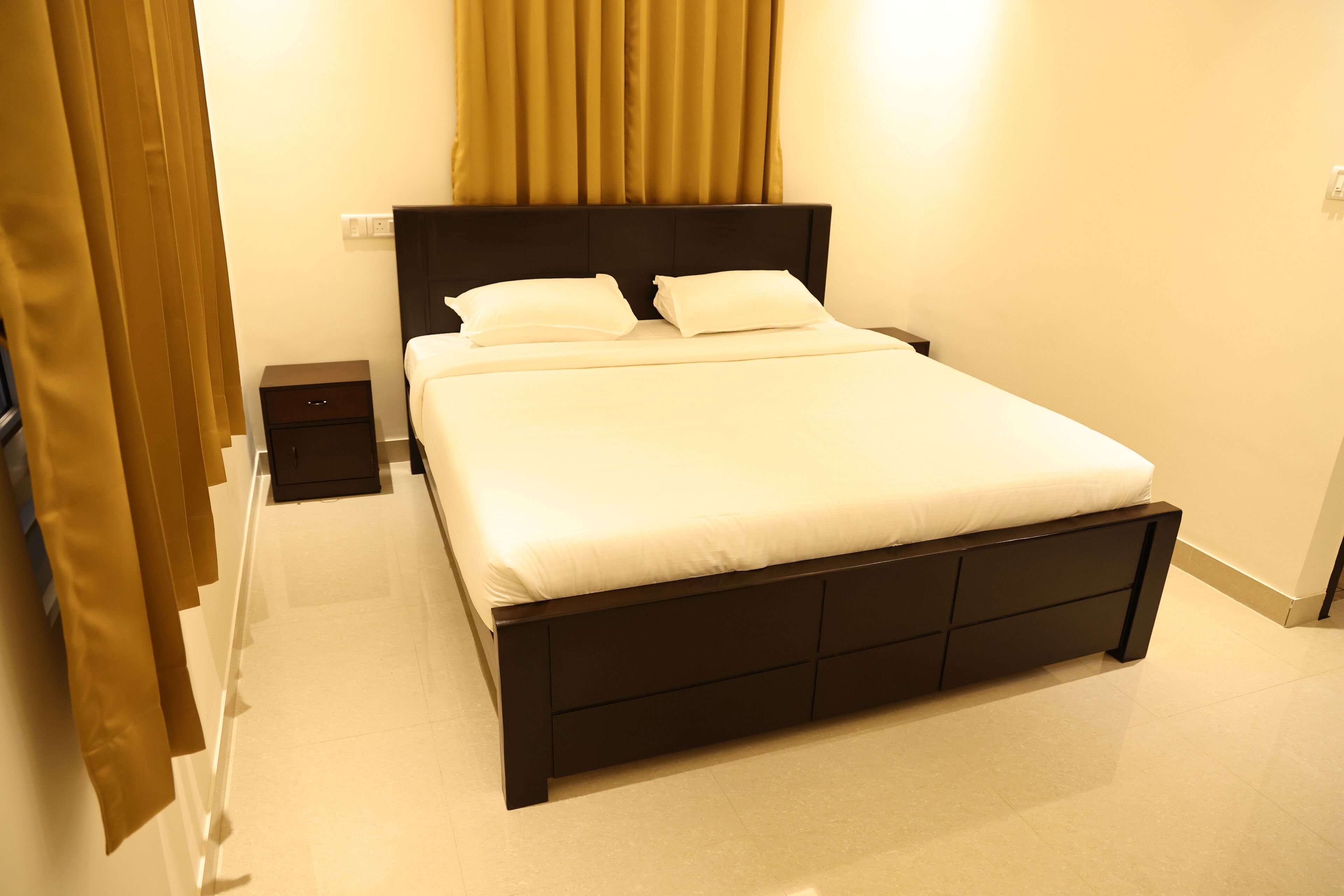 Uma cama bem arrumada com cabeceira escura e roupa de cama branca, uma pequena mesa de cabeceira e uma cortina à esquerda.