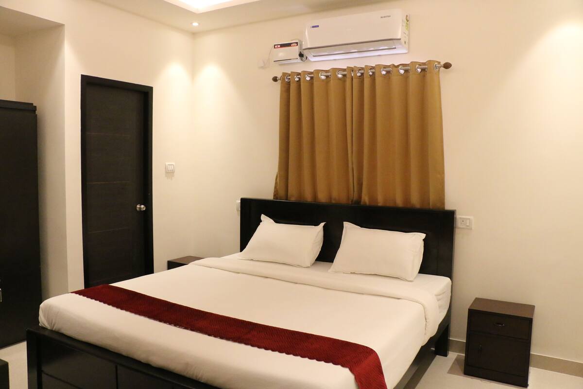 Cama bem arrumada com roupa de cama branca e manta vermelha, cabeceira escura e cortina marrom.