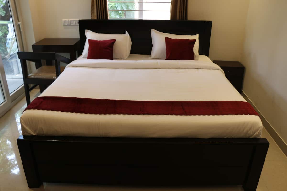 Uma cama grande com lençóis brancos e almofadas vermelhas, uma mesa pequena e uma cadeira num quarto com janela.