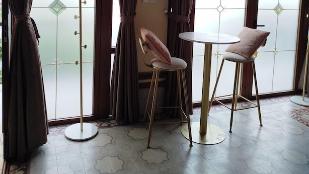 Um quarto com duas janelas em arco, uma mesa redonda com duas cadeiras, um objeto decorativo na parede e um piso estampado.