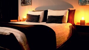 Uma cama com edredom branco, duas almofadas pretas e cabeceira de madeira. Há duas mesas de cabeceira com velas.