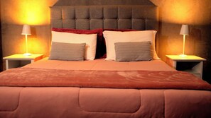 Uma cama com cabeceira estofada, duas mesas de cabeceira com abajures e um cobertor vermelho.