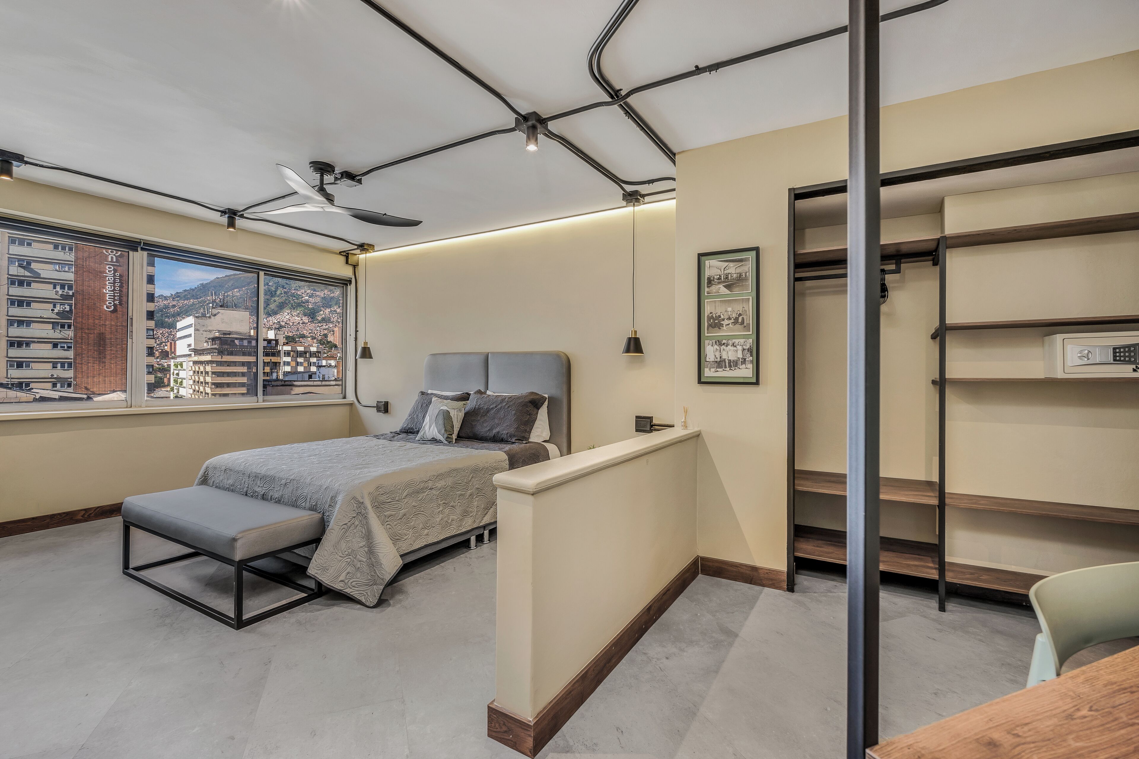 Quarto com cama, um banco e vista para edifícios através de grandes janelas.