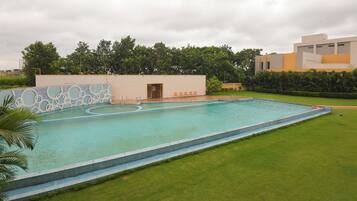 Uma piscina com design geométrico em azulejos, rodeada por um relvado bem cuidado e um edifício com fachada moderna.