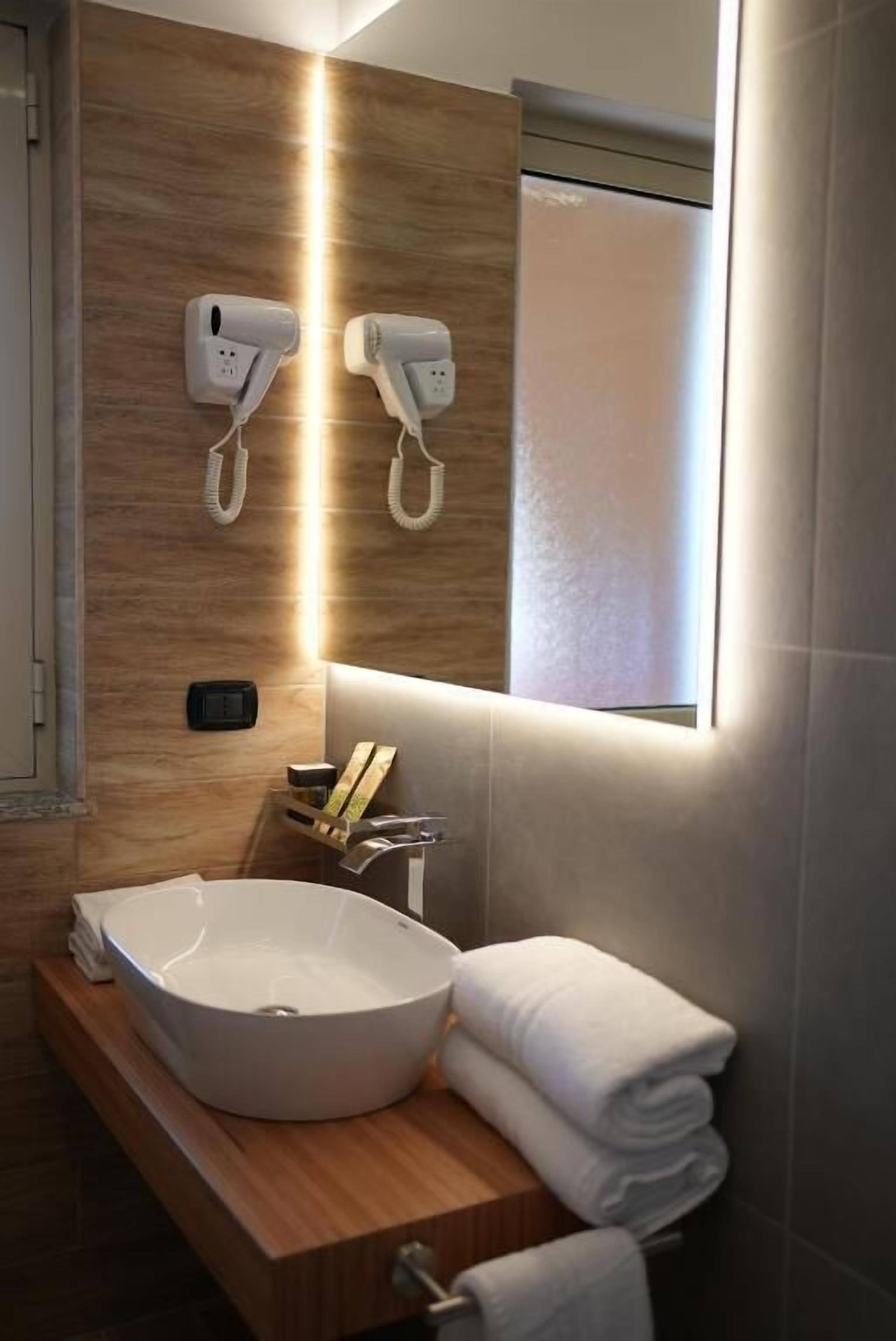 Um banheiro moderno com uma pia grande e branca, móvel de vaidade em madeira e dois secadores de cabelo fixados na parede.