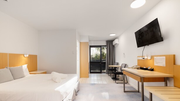 Quarto de hotel com cama, escrivaninha, cadeira, televisão e janela com cortinas.