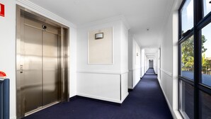Corredor de hotel com carpete azul, luminária na parede e elevador.