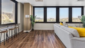 Uma sala de estar moderna com uma janela ampla, um sofá cinza com almofadas amarelas, piso de madeira e um espaço bar com banquetas.