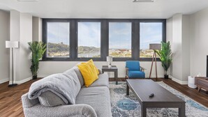 Uma sala de estar moderna com um sofá cinza, uma poltrona azul, uma mesa de centro e grandes janelas com vista para a cidade.