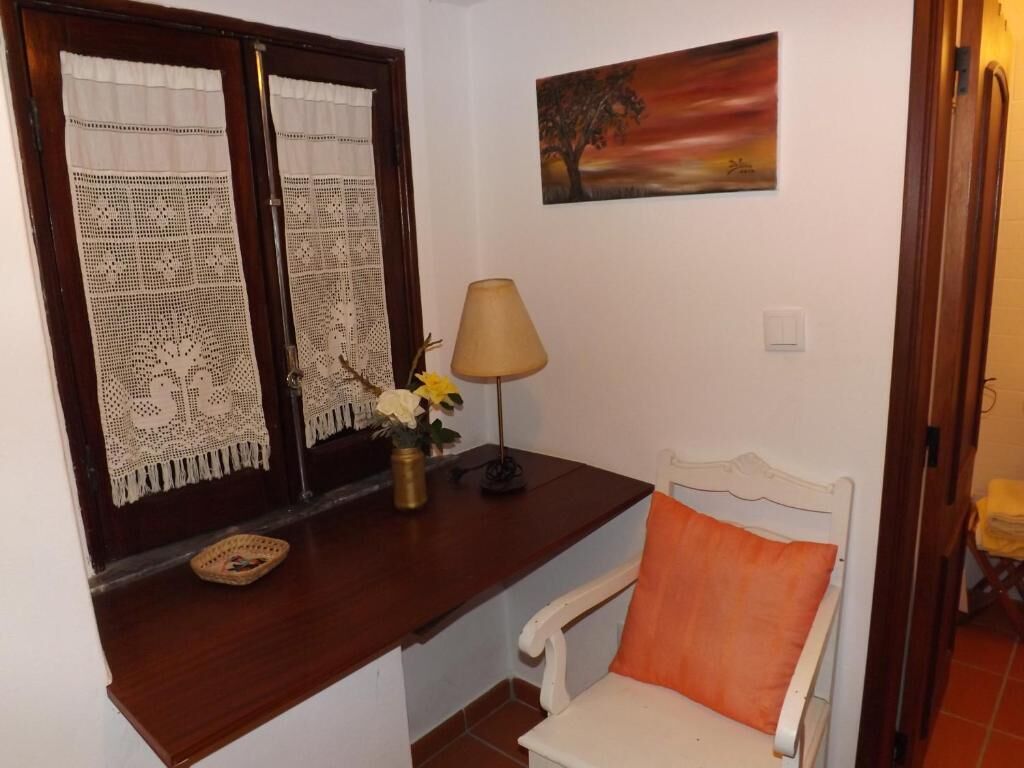 Quarto com escrivaninha de madeira, cadeira branca com almofada laranja, luminária e pintura de árvore na parede.