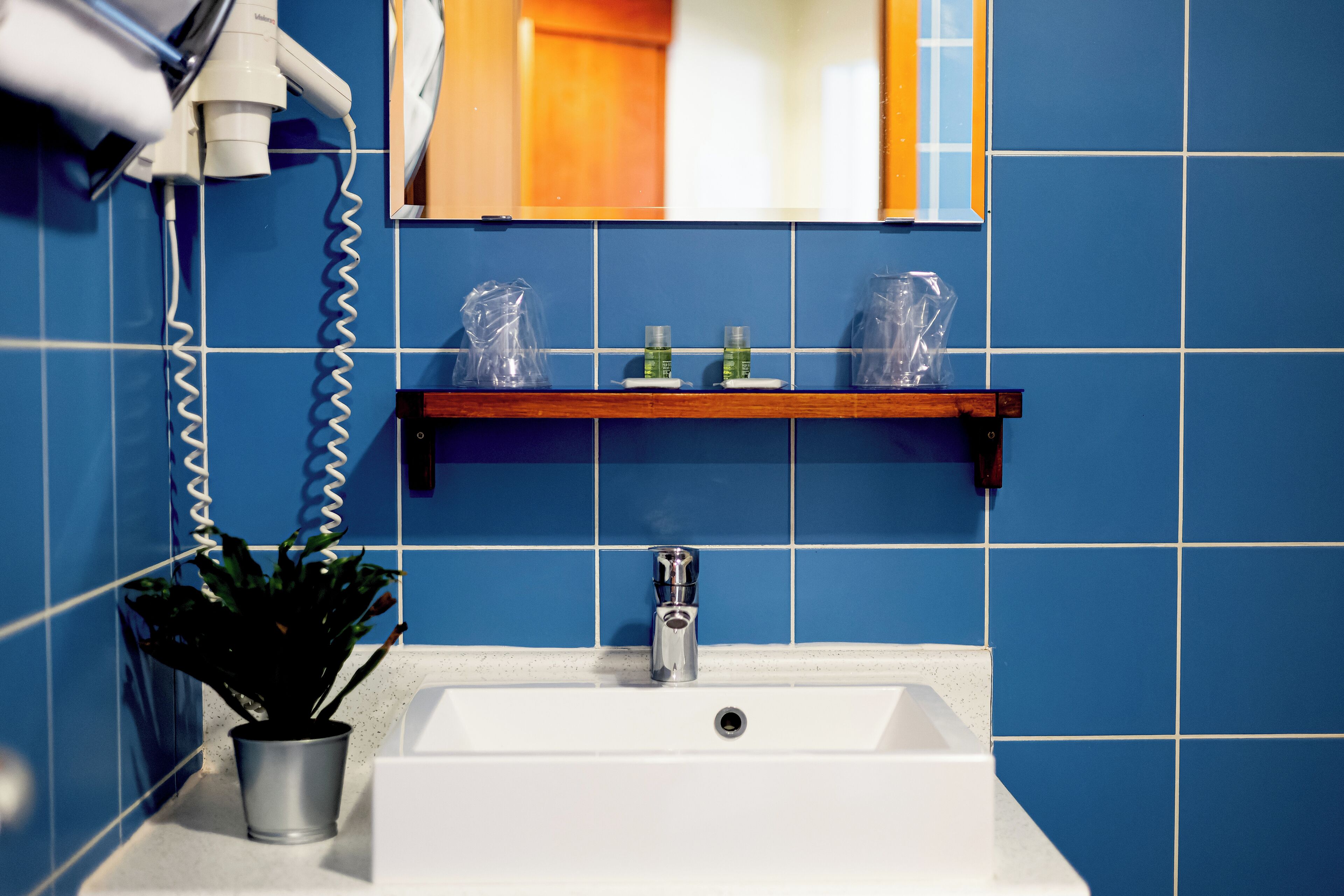 Casa de banho com azulejos azuis, uma pia branca, um espelho, um secador de cabelo e uma estante com artigos de higiene.