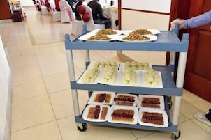Carrinho de comida com bandejas de diversos pratos, incluindo aparentemente doces e sobremesas, em um espaço com piso ladrilhado e uma porta ao fundo.