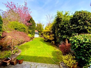 Um jardim bem cuidado com gramado, arbustos variados e uma pequena construção ao longe.