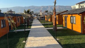 Uma fileira de cabanas laranjas com um caminho pavimentado que leva a um portão, cercada por vegetação e montanhas ao fundo.