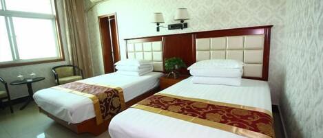 Quarto de hotel com duas camas, uma mesinha, uma cadeira, uma janela e uma luminária na parede.