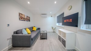 Uma sala de estar moderna com um sofá cinzento, uma mesa de centro, uma TV fixada na parede e uma pequena mesa lateral.