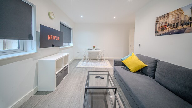 Uma sala de estar moderna com um sofá cinzento, uma mesa de centro de vidro, um rack de televisão branco e uma área de jantar com mesa e cadeiras brancas.