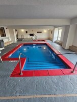 Piscina interior retangular com borda em azulejos vermelhos e azuis, uma escada de metal e chão revestido de azulejos.