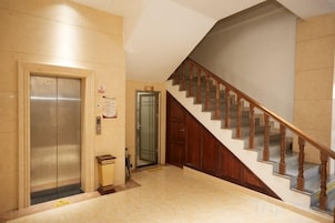Um saguão de hotel com piso de mármore, um elevador, uma escada com corrimãos de madeira e uma porta com painel de vidro.
