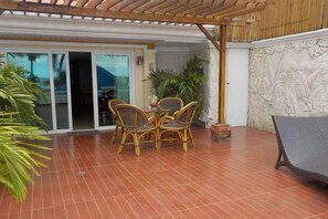 Um pátio externo com mesa e cadeiras, uma área de estar coberta e uma porta de vidro deslizante que dá para uma entrada coberta.