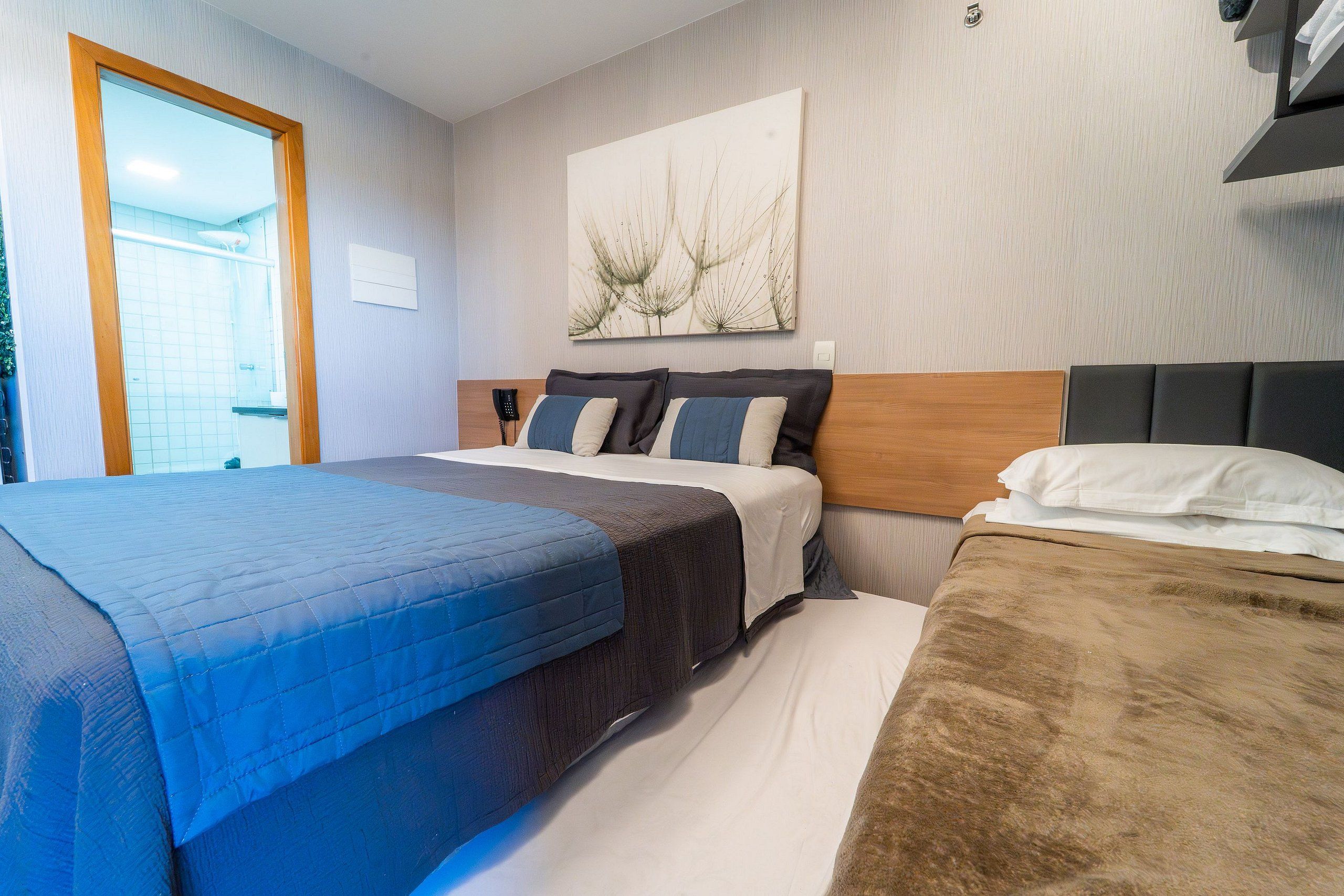 Quarto de hotel com uma cama grande, uma cama menor, uma cabeceira de madeira, uma obra de arte na parede e uma porta que dá para um espaço com chuveiro.
