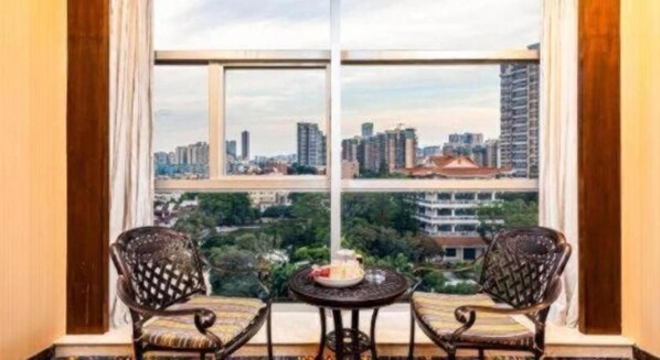 Quarto com duas cadeiras de vime, uma mesa redonda pequena, uma grande janela com vista para a cidade e uma cortina à esquerda.