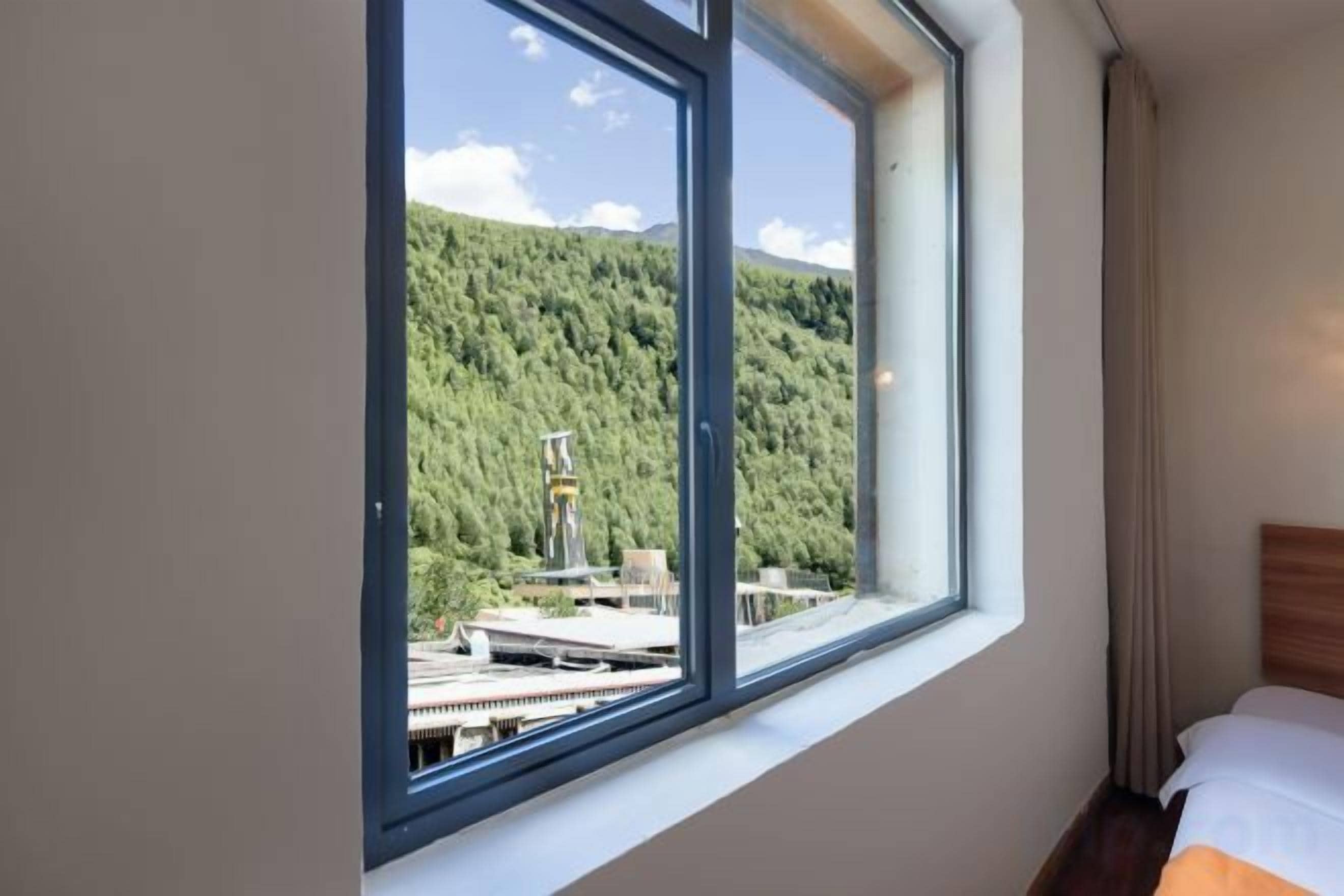 Quarto de hotel com uma grande janela, uma cama e vista para uma área arborizada.