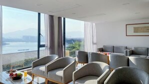 Quarto de hotel moderno com uma grande janela que oferece vista para um corpo de água e montanhas. Há várias cadeiras estofadas em cinza e uma pequena mesa com uma cesta de frutas e um copo de suco de laranja.