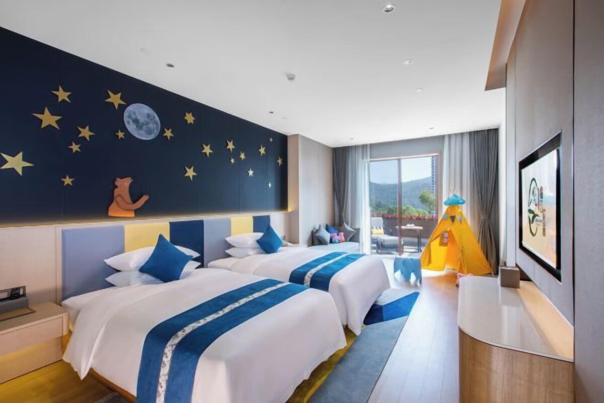 Quarto de hotel com duas camas, uma escrivaninha, uma cadeira, janela com vista e decoração de estrelas e luas na parede.