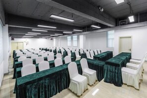 Uma sala de conferências com fileiras de mesas e cadeiras, teto alto com iluminação industrial e uma porta ao fundo.