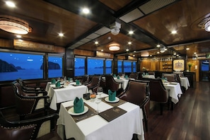 Restaurante com mesas preparadas para refeições, grandes janelas com vista para o mar e teto de madeira.