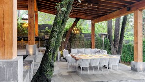 Um pátio ao ar livre com pérgola de madeira, piso de pedra e uma mesa redonda com cadeiras brancas.