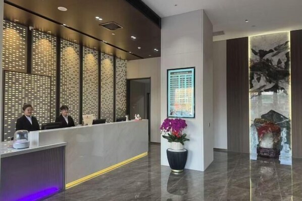 Uma área de recepção de hotel moderna, com balcão de atendimento, dois funcionários, painéis decorativos na parede, uma obra de arte emoldurada e um vaso com planta.