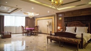 Quarto de hotel com cama, um banco, uma escrivaninha com cadeiras, um candelabro e uma janela grande com cortinas.