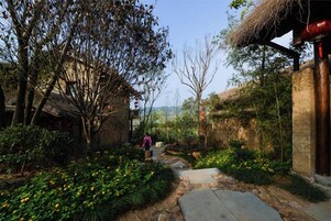Um caminho de jardim com um banco de pedra, rodeado por vegetação e uma estrutura com telhado de palha.