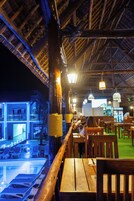 Um restaurante em ambiente interno com mesas e cadeiras de madeira, área de piscina e telhado de palha.