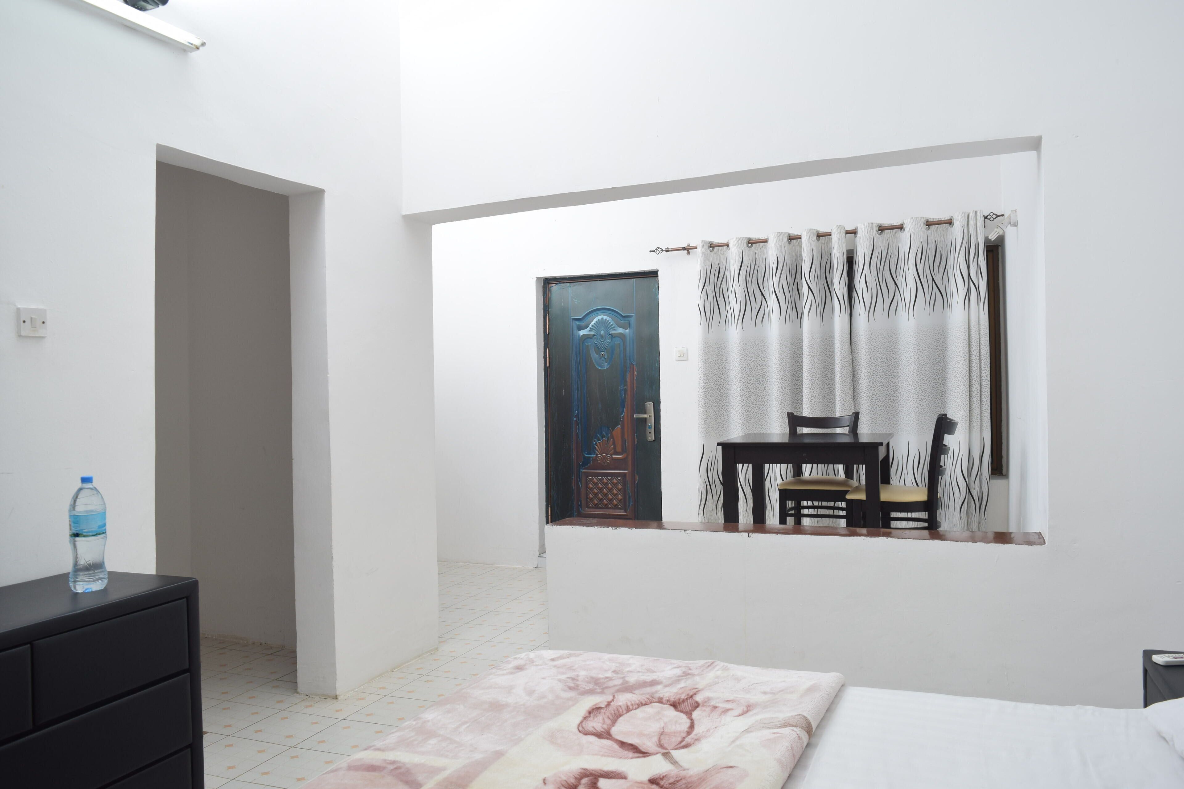 Quarto de hotel com cama, cômoda, uma garrafa de água na cômoda, janela com cortinas e porta com detalhes decorativos.