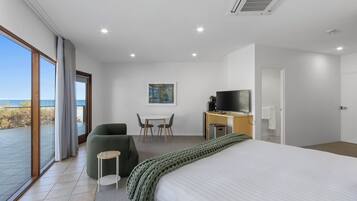 Quarto de hotel moderno com uma cama grande, uma mesa de escritório com televisão, área de estar com cadeiras e vista para a praia através da porta de vidro.