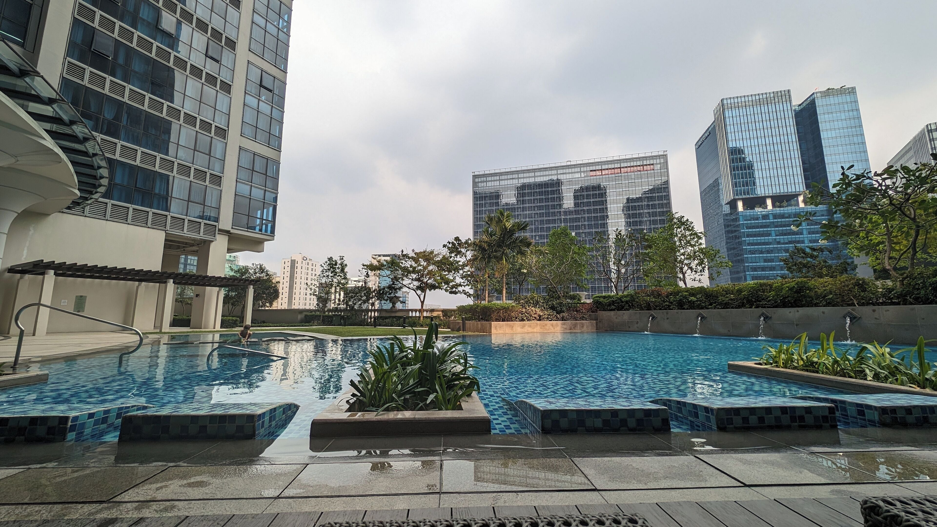 Piscina com bordas azulejadas, cercada por vegetação e edifícios modernos.