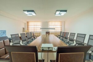 Uma sala de conferências com uma mesa comprida, cadeiras, um projetor e janelas com cortinas.