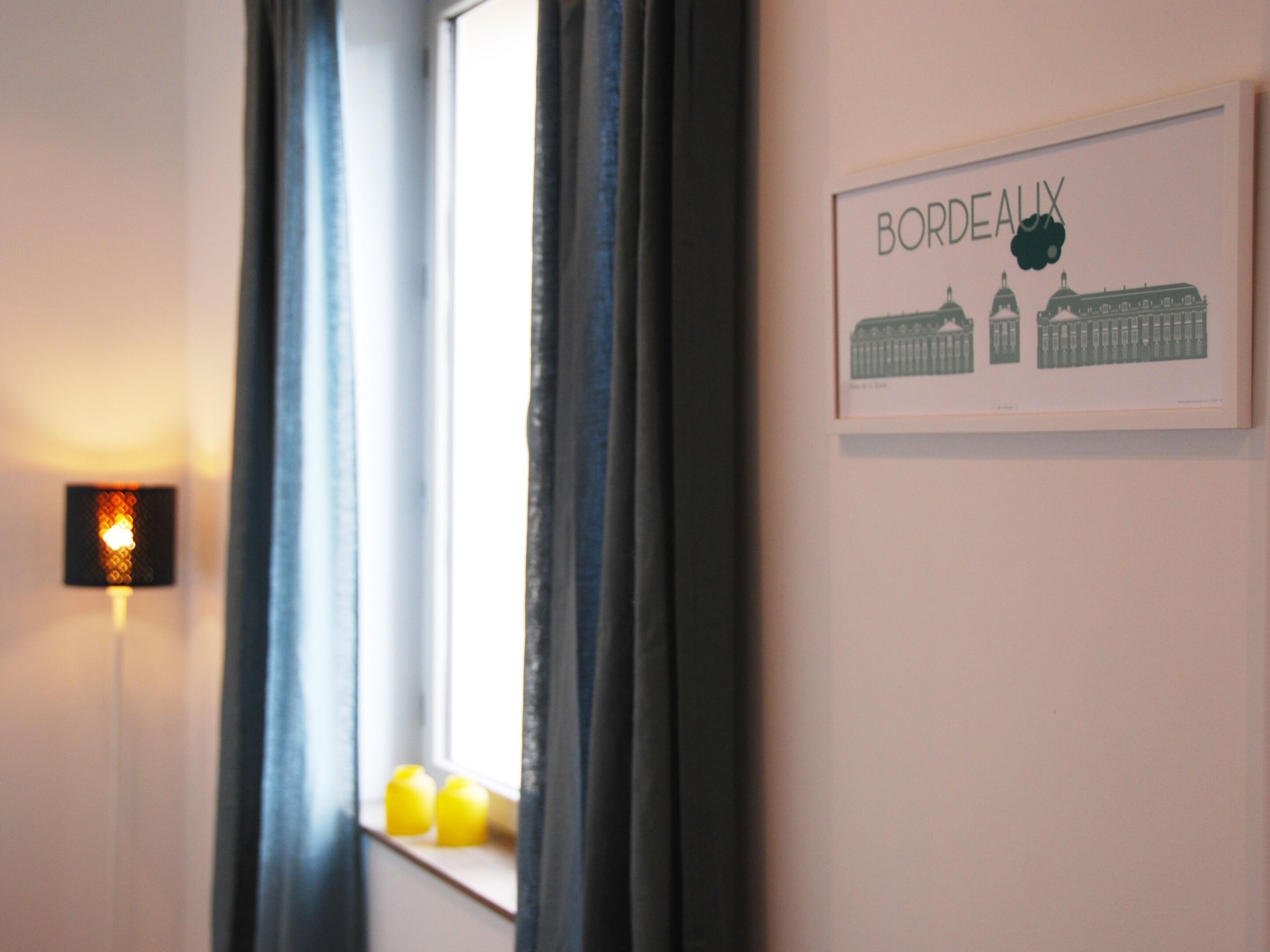 Quadro emoldurado de Bordeaux numa parede, janela com cortinas e luminária fixada na parede.