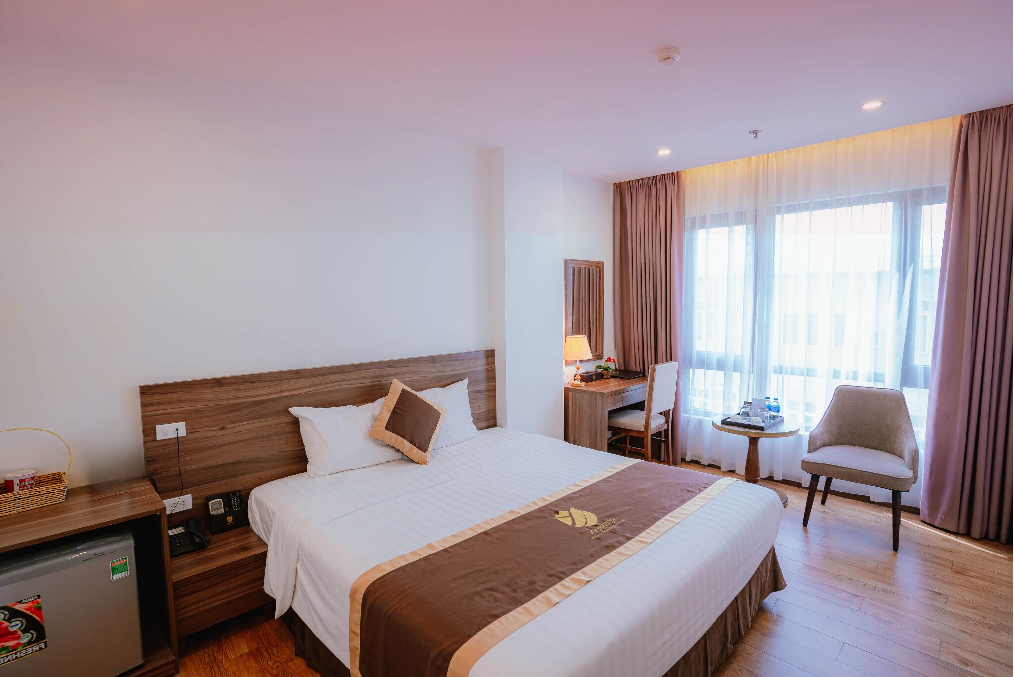 Quarto de hotel com uma cama grande, escrivaninha, cadeira, mesa pequena e geladeira.