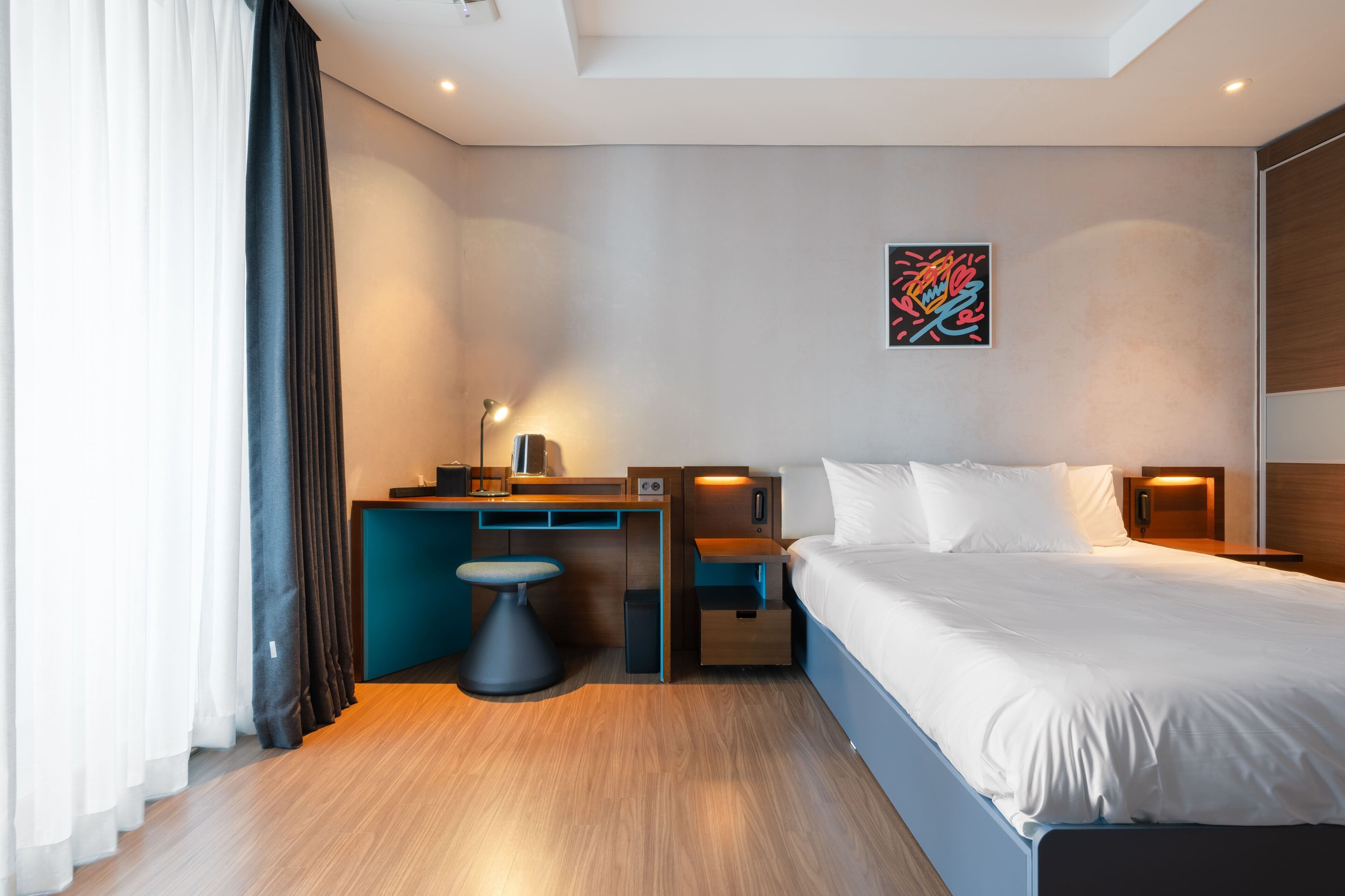 Quarto de hotel moderno com cama, escrivaninha, cadeira e um banquinho.
