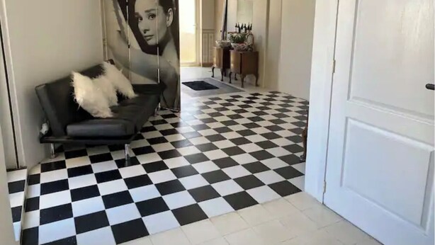 Um corredor com chão quadriculado em preto e branco, um sofá preto com almofada branca, e um pôster grande na parede.