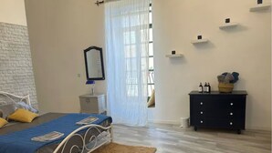 Quarto com cama, mesa de cabeceira, cômoda e janela com cortinas leves.