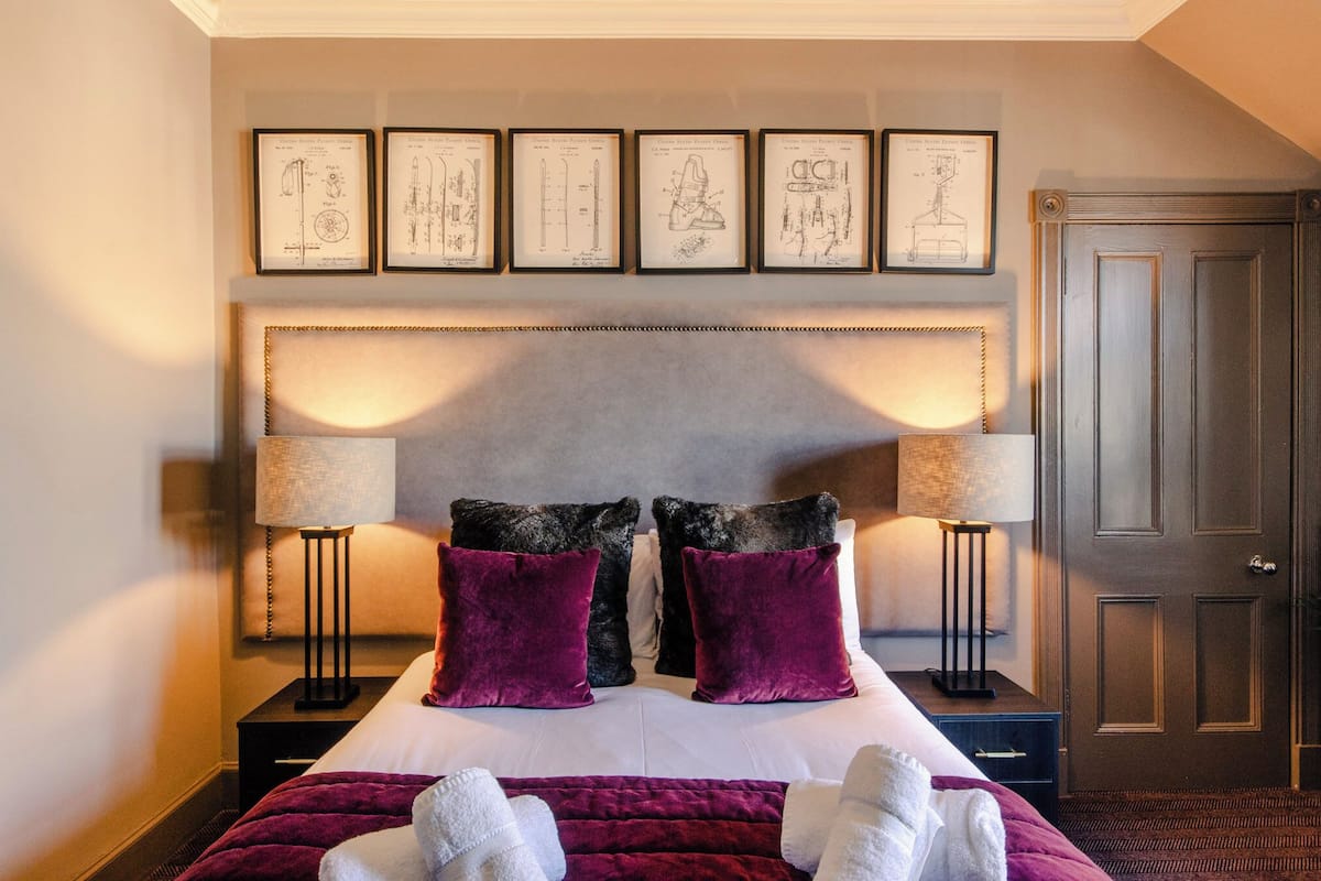 Quarto de hotel com uma cama grande, duas mesas de cabeceira com luminárias, cabeceira com patentes emolduradas e uma porta.