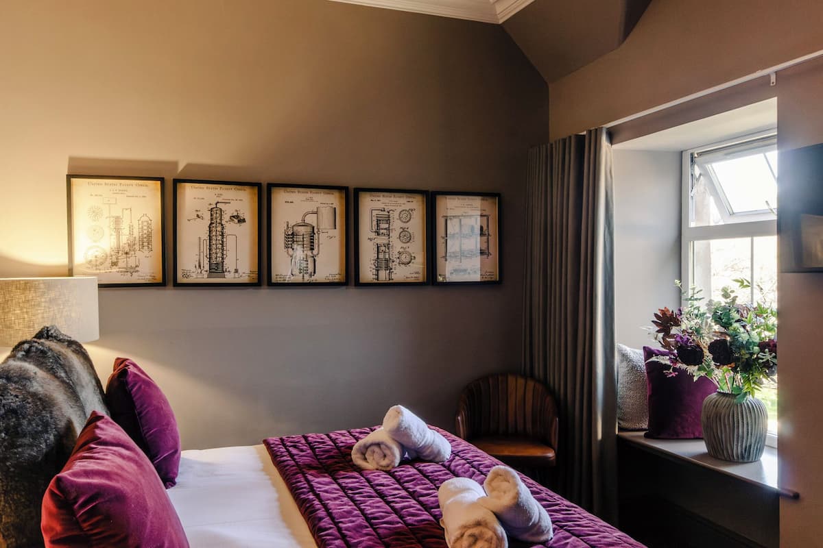 Quarto de hotel com cama, almofadas, uma cadeira, janela com cortinas e quatro desenhos de patentes emoldurados na parede.